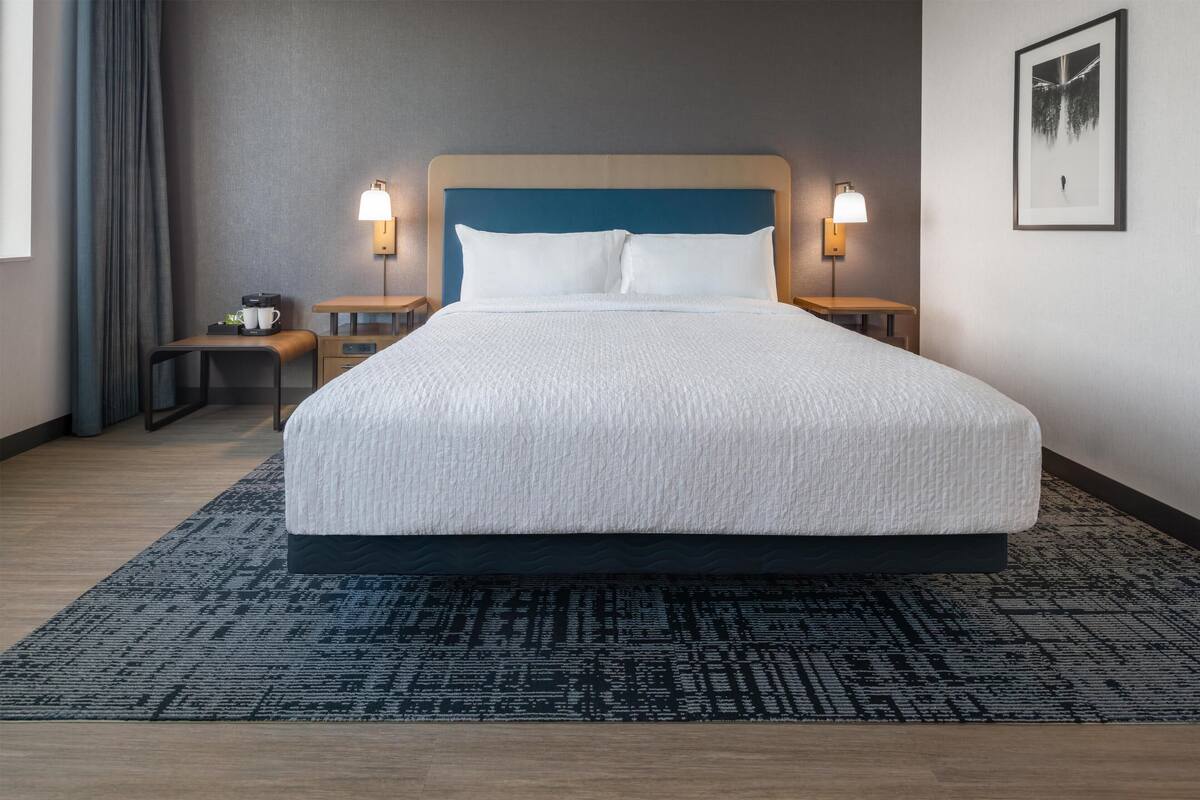 Quarto de hotel com uma cama grande, mesas de cabeceira, abajur, um quadro na parede e uma janela com cortinas.