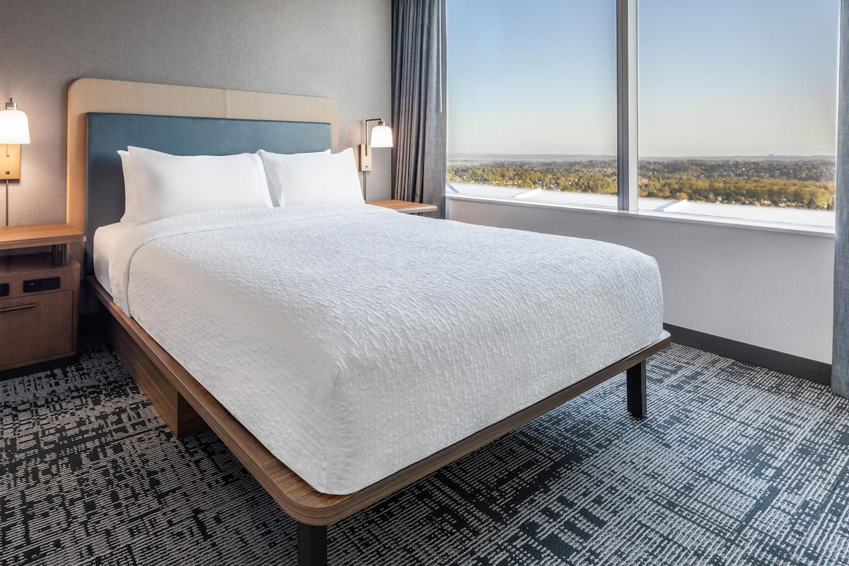 Quarto de hotel com uma cama grande, mesa de cabeceira, abajur e vista para a cidade pela janela.