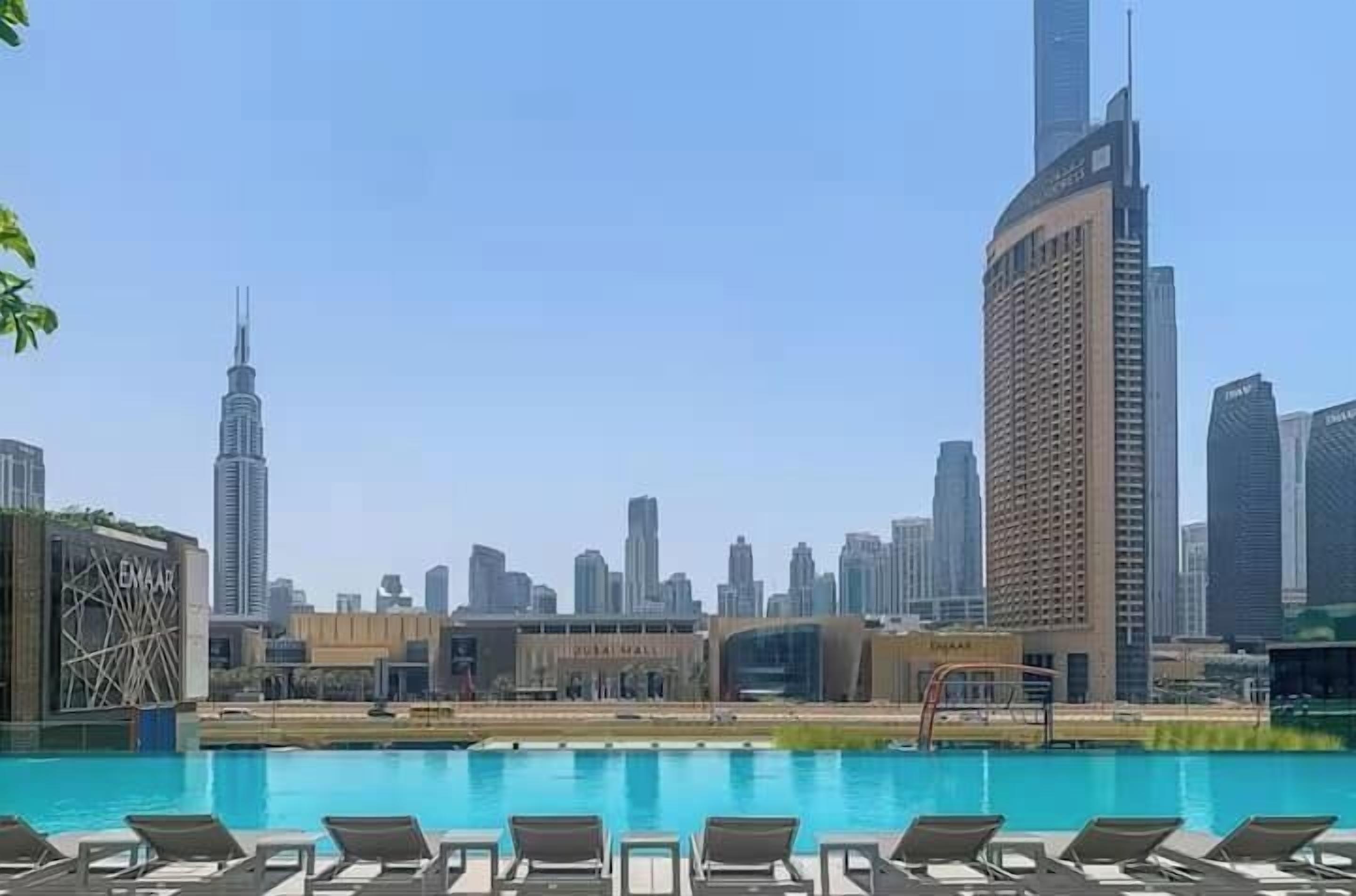 Vista da piscina com espreguiçadeiras, um edifício moderno com fachada singular e um horizonte urbano ao fundo.