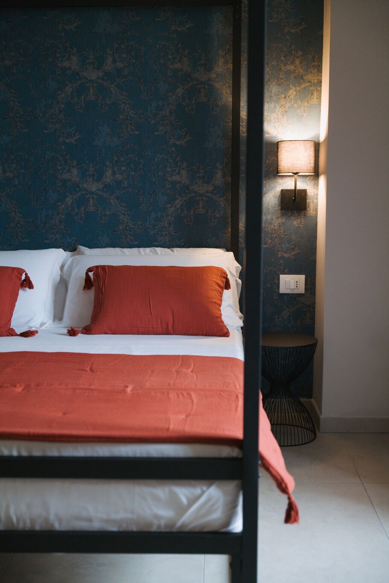Cama com roupa de cama branca e vermelha, parede com padrão azul e luminária fixada na parede.
