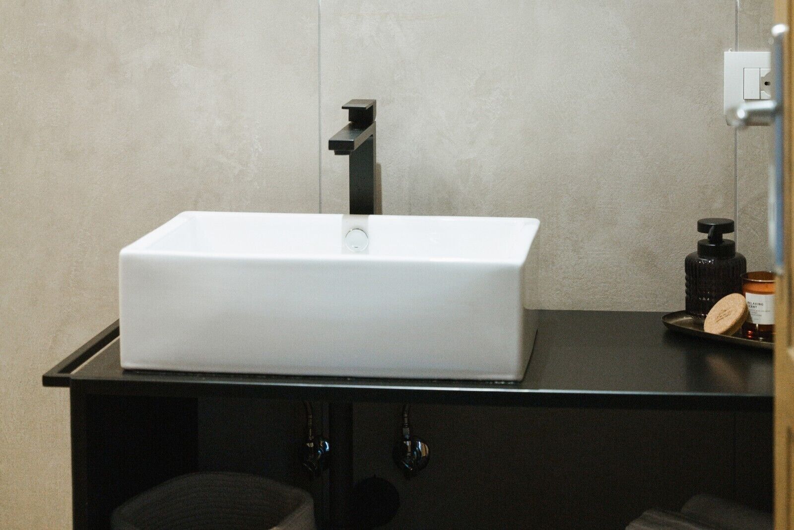 Um banheiro com uma pia retangular branca, uma torneira preta e um pequeno dispensador de sabonete preto no balcão.