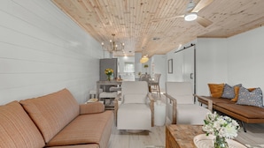 Uma sala de estar moderna com teto de madeira, um sofá, poltronas, uma mesa de jantar e uma cozinha ao fundo.