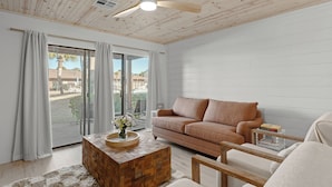 Sala de estar com teto de madeira, sofá bege, mesa de centro de madeira e vista para uma piscina e edifícios lá fora.