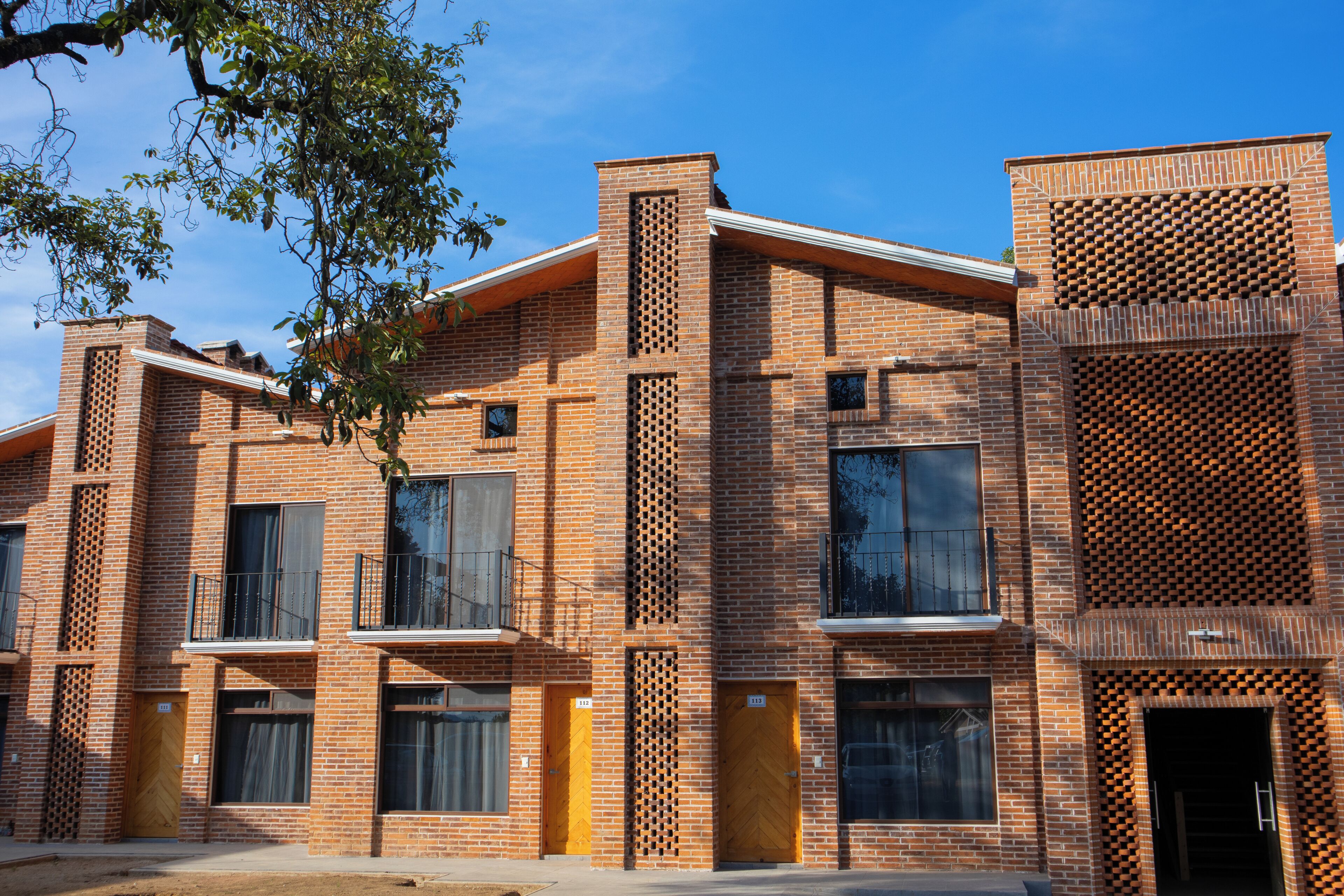 Um edifício de vários andares em tijolo, com varandas, janelas amplas e uma árvore em primeiro plano.