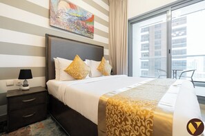 Quarto de hotel com uma cama grande, mesa de cabeceira com abajur, quadro na parede e vista da cidade através de grandes janelas.
