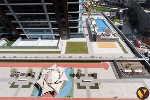Um edifício moderno com piscina no terraço, praça arborizada e instalação artística geométrica.