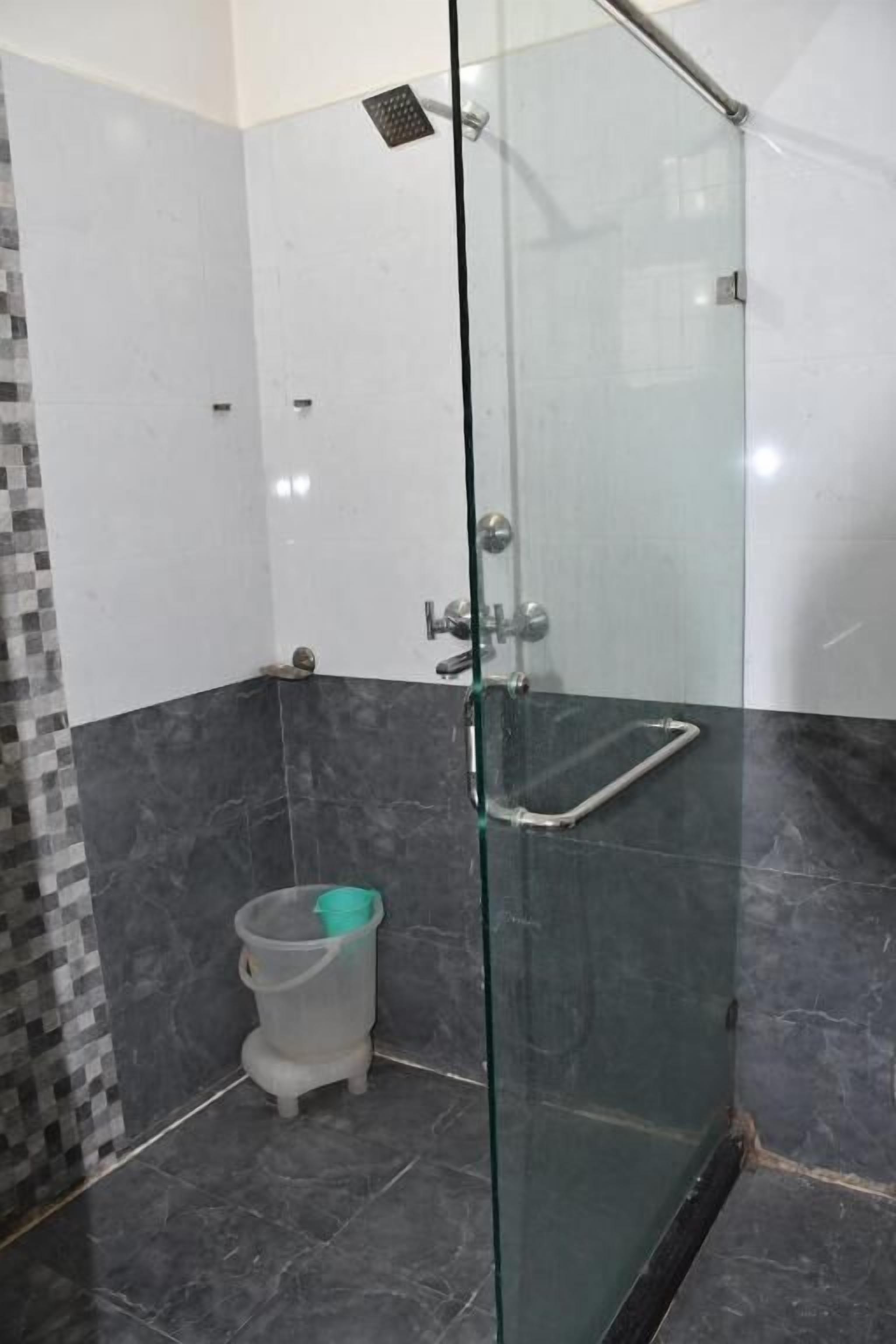Um chuveiro com divisória de vidro, corrimão metálico, uma pequena lixeira branca e piso cerâmico.