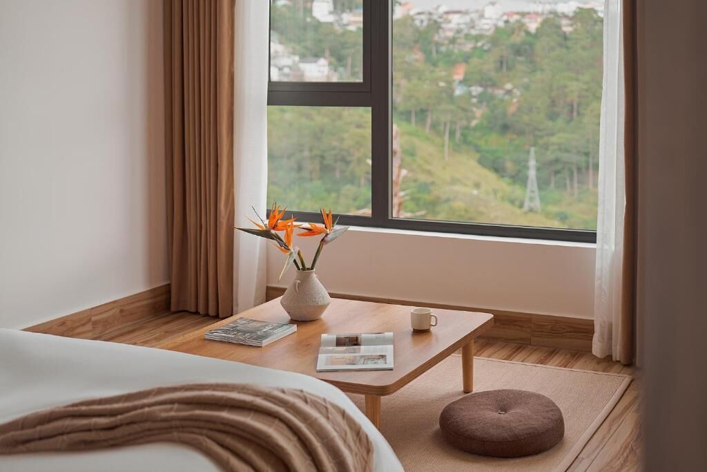 Quarto com cama, mesa de cabeceira com um vaso de flores, uma revista e janela com vista para o verde.