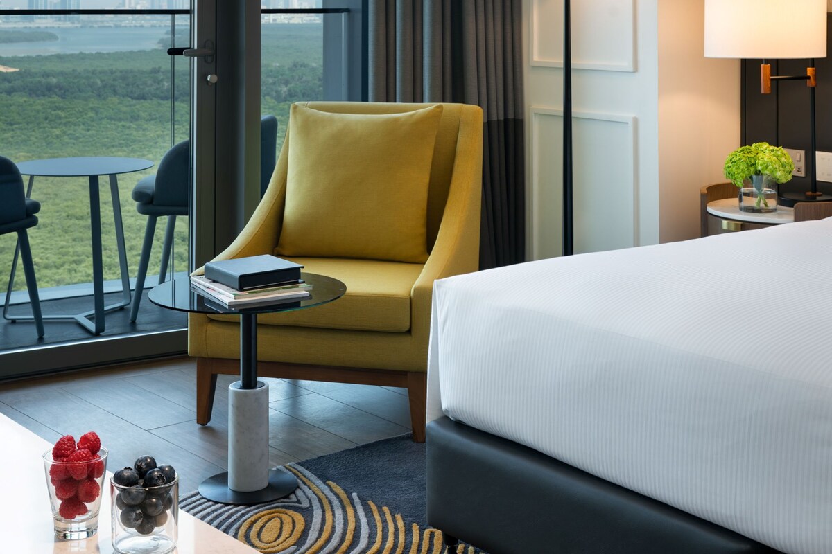 Quarto de hotel com uma cama, uma poltrona amarela, uma mesa pequena, uma luminária e vista para a cidade através de grandes janelas.