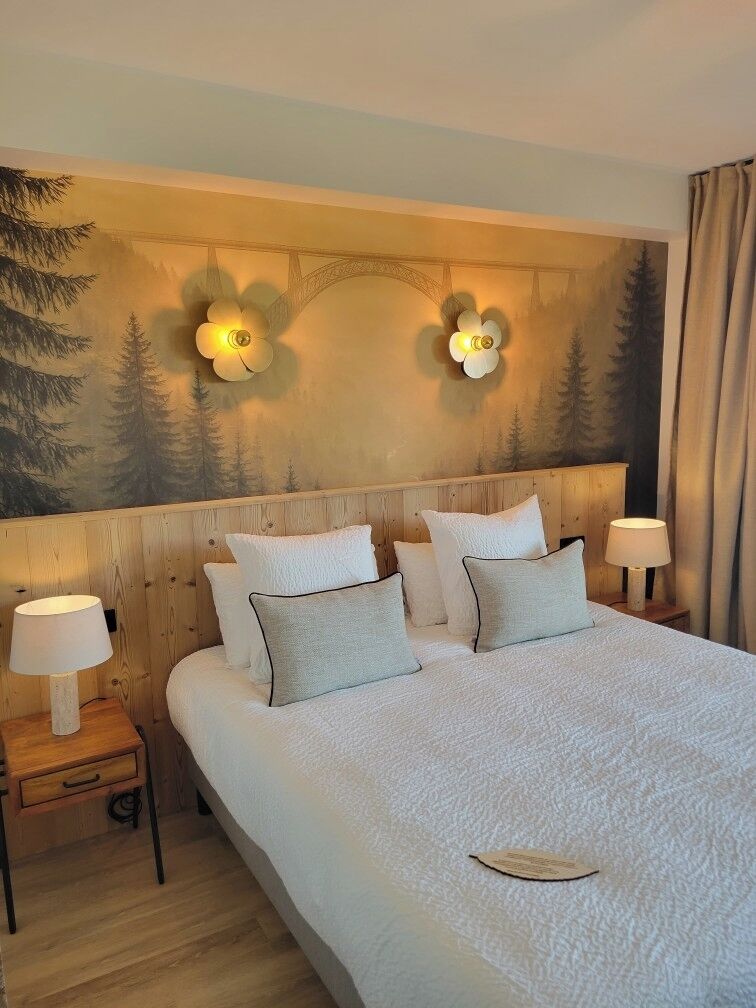 Uma cama grande com roupa de cama branca, duas luminárias de cabeceira e um mural na parede com a cena de uma floresta.