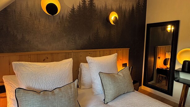 Um quarto com teto inclinado, cama com roupa de cama branca, e um mural com árvores e uma lua.