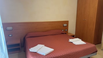 Uma cama de solteiro com colcha vermelha estampada, cabeceira de madeira, mesa de cabeceira, guarda-roupa de madeira e uma tomada na parede.