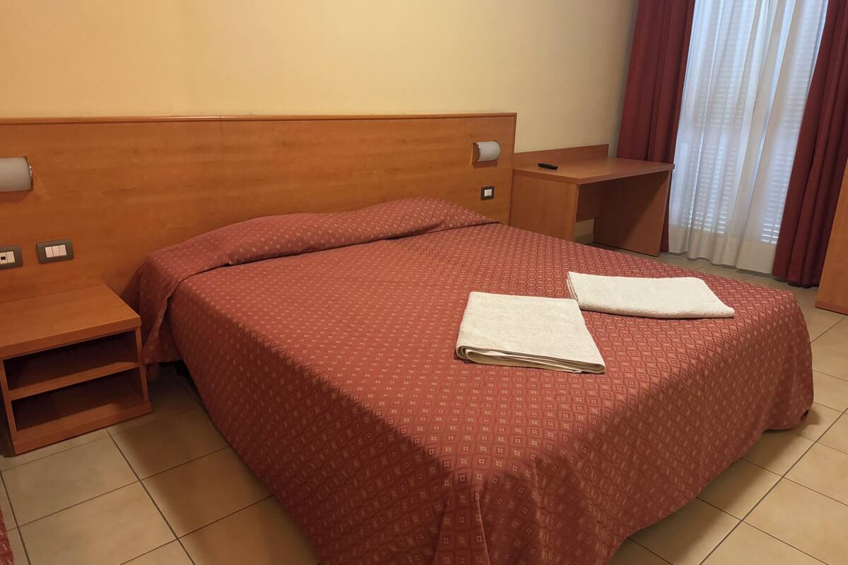 Quarto de hotel com cabeceira de madeira, cama com colcha estampada em vermelho, duas toalhas sobre a cama, mesa de cabeceira e janela com cortinas.