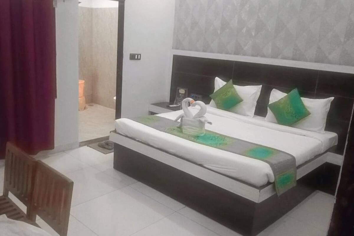 Quarto de hotel com uma cama, uma cadeira de madeira, uma mesinha de cabeceira e um banheiro visível através de uma porta aberta.