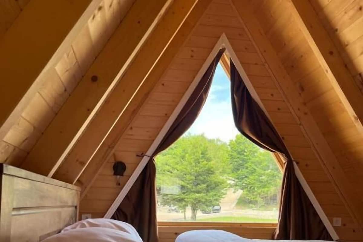 Um quarto de sótão aconchegante, com teto inclinado de madeira, uma cama de solteiro com roupa de cama branca e uma janela com vista para uma paisagem verde.