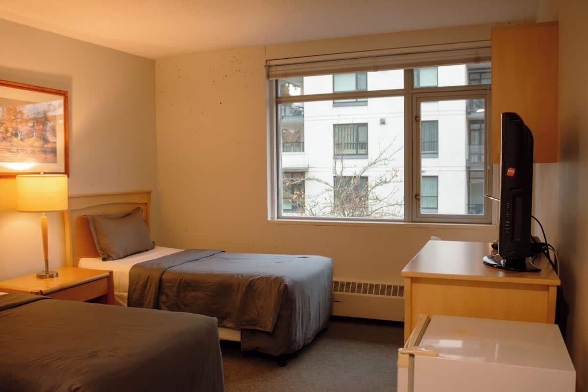 Quarto de hotel com duas camas, uma escrivaninha com computador, uma janela com vista para edifícios e uma luminária.