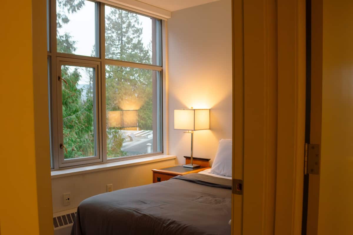 Quarto de hotel com uma cama, mesa de cabeceira, luminária e janela com vista para árvores.
