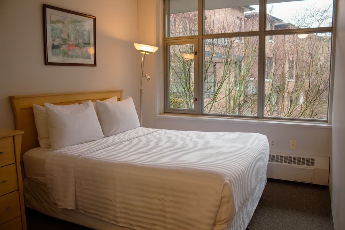 Uma cama bem arrumada com roupa de cama branca, uma luminária de cabeceira e uma janela com vista para árvores e edifícios.