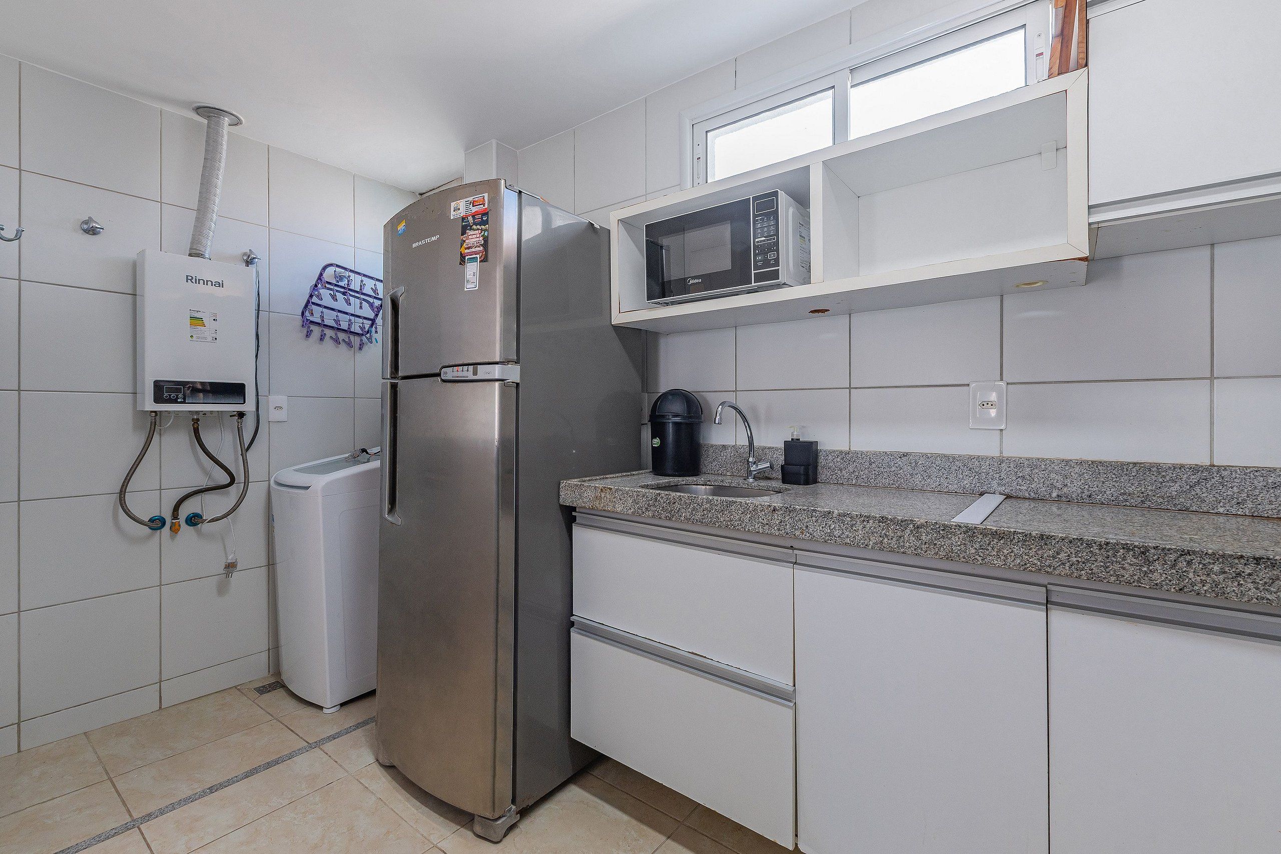 Uma cozinha moderna com armários brancos, uma geladeira de aço inoxidável, micro-ondas embutido e aquecedor de água.