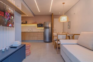 Cozinha moderna com frigorífico, micro-ondas e armários de madeira. Há uma área de refeições com mesa e cadeiras, e um sofá na sala de estar.