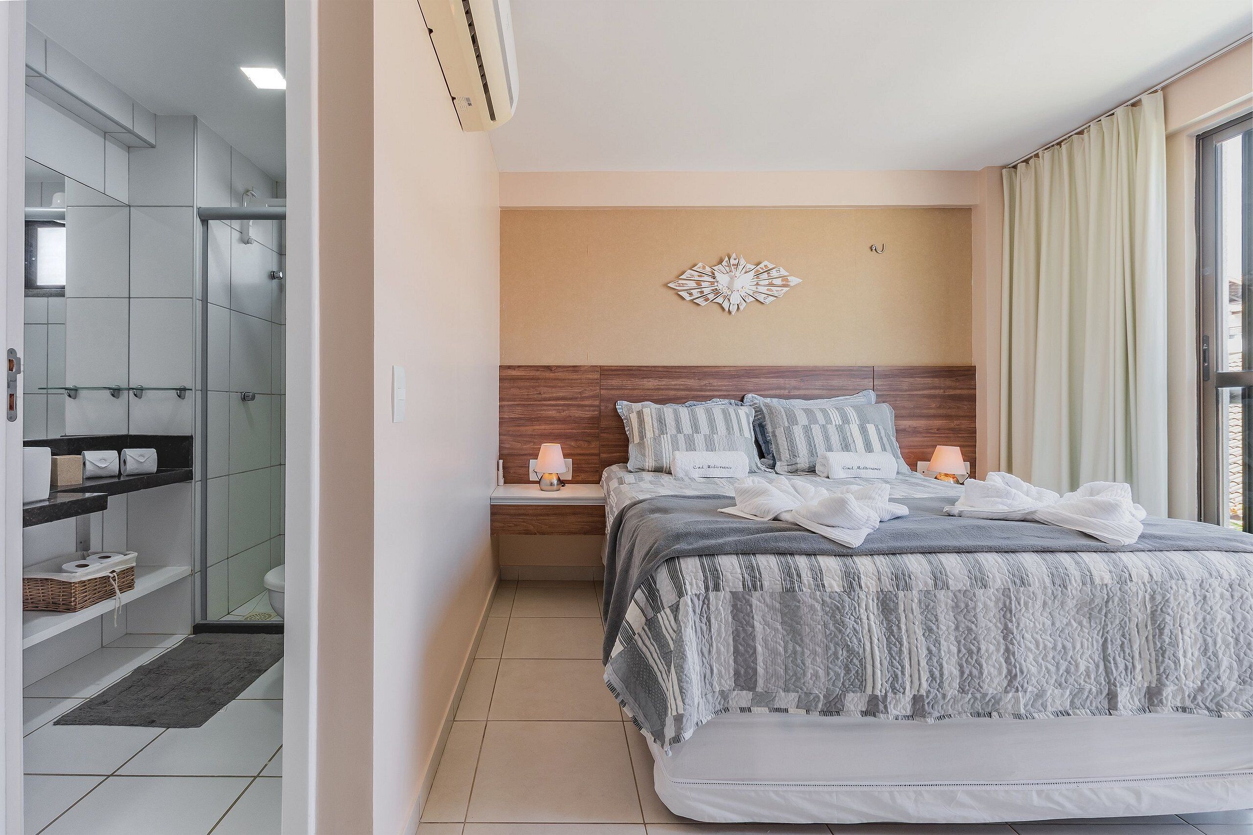 Quarto de hotel com cama, mesa de cabeceira, objeto decorativo na parede e banheiro com chuveiro e vaso sanitário.