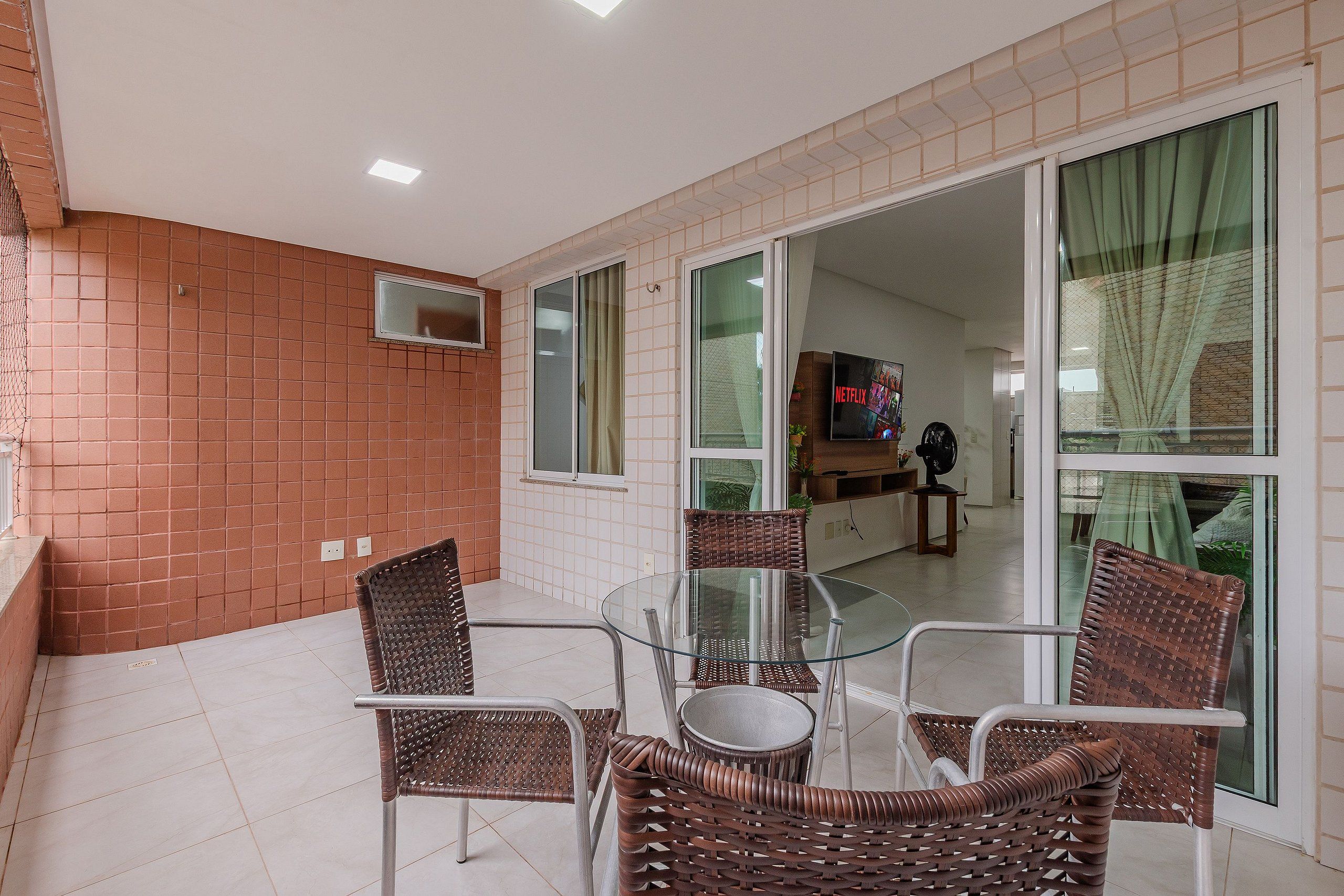 Uma varanda com mesa de vidro, cadeiras de vime, piso de azulejos e uma TV ao fundo.