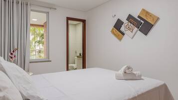 Quarto de hotel com cama, janela com cortinas, banheiro visível pela porta e parede com quadros emoldurados.