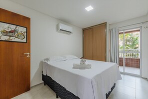 Quarto de hotel com cama, ar condicionado fixado na parede, porta e janela com cortinas.
