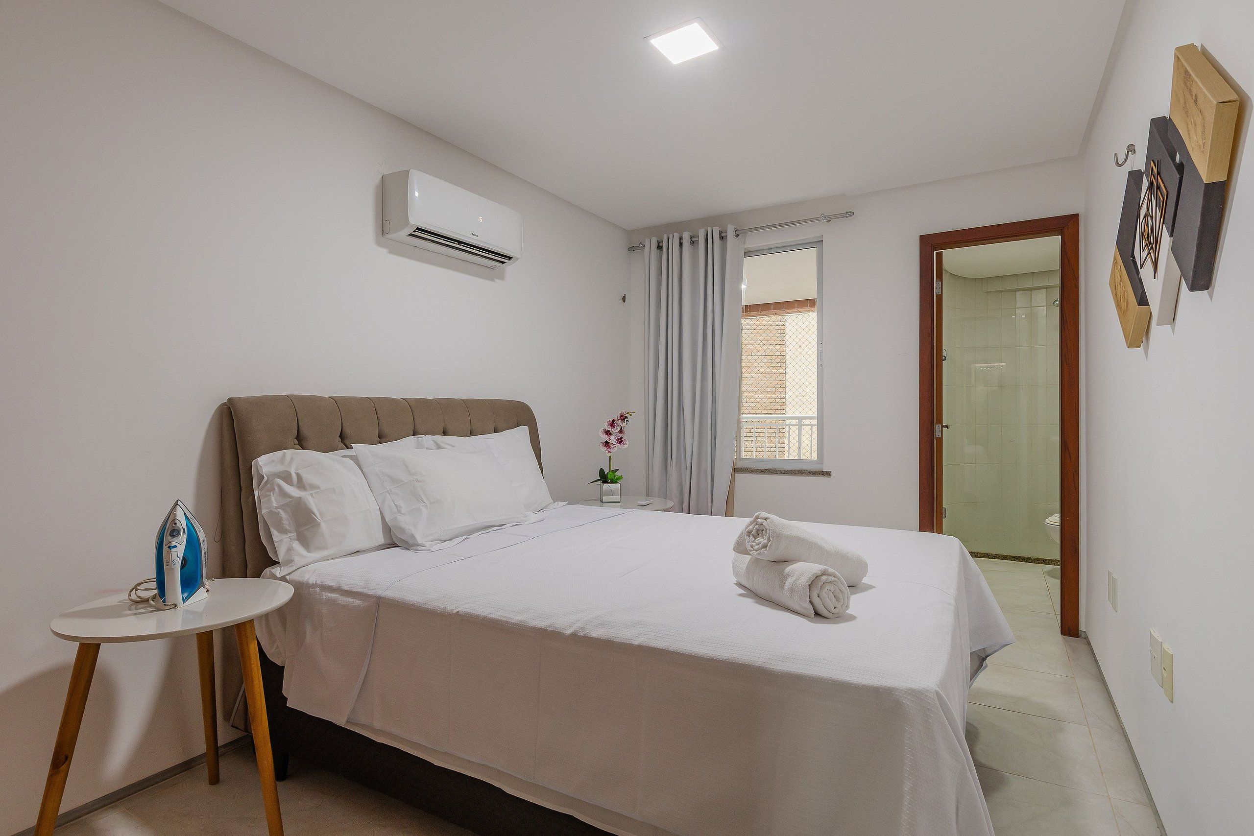 Uma cama bem arrumada com lençóis brancos, uma pequena mesa de cabeceira com um ferro e uma janela com cortinas.