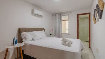 Uma cama bem arrumada com lençóis brancos, uma pequena mesa de cabeceira com um ferro e uma janela com cortinas.