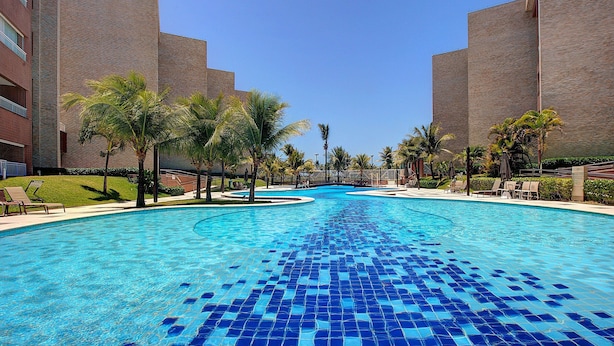 Uma piscina com azulejos de mosaico azul, rodeada por palmeiras e um edifício de tijolos.