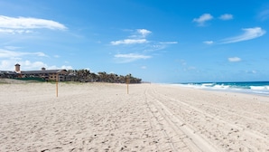 Uma extensa praia de areia com pegadas que levam ao oceano, um edifício com uma torre e um céu azul límpido.
