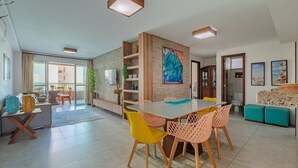 Ampla sala de estar com mesa de jantar, cadeiras coloridas, sofá e cozinha ao fundo.