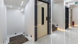 Um elevador moderno com porta bege e detalhes em preto, parede branca e piso de azulejos brilhantes.