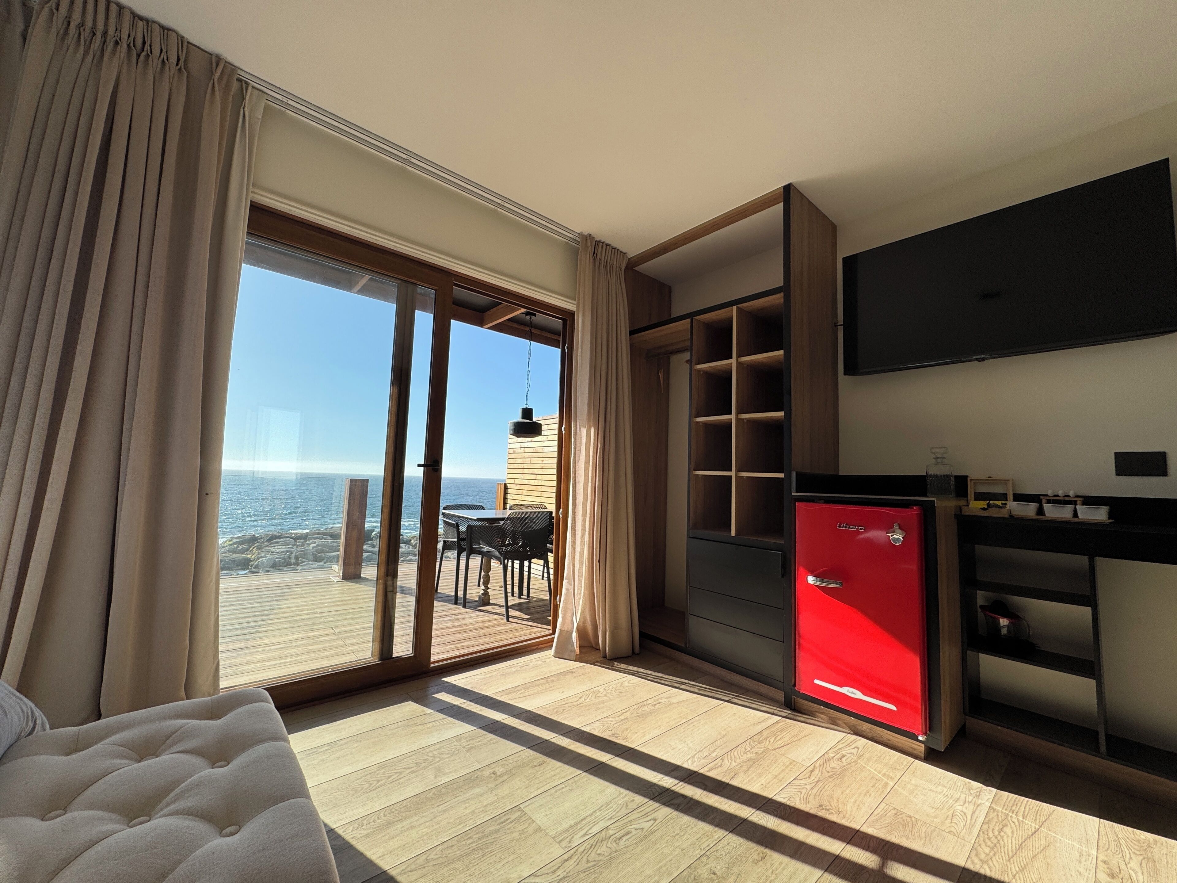 Quarto moderno com porta de vidro deslizante que dá para uma varanda, um minibar vermelho e uma TV de tela plana fixada na parede.