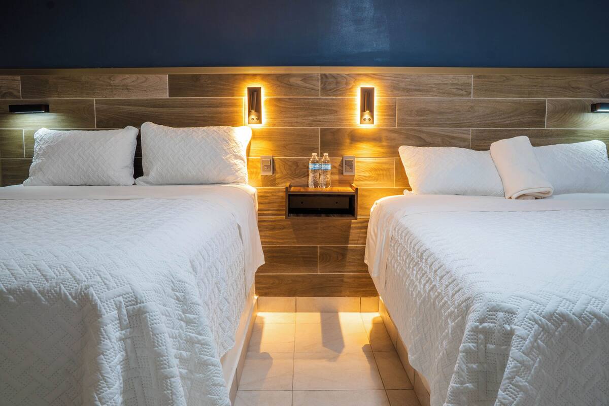 Quarto de hotel com duas camas, cabeceira de madeira, mesas de cabeceira e luminárias fixadas na parede.