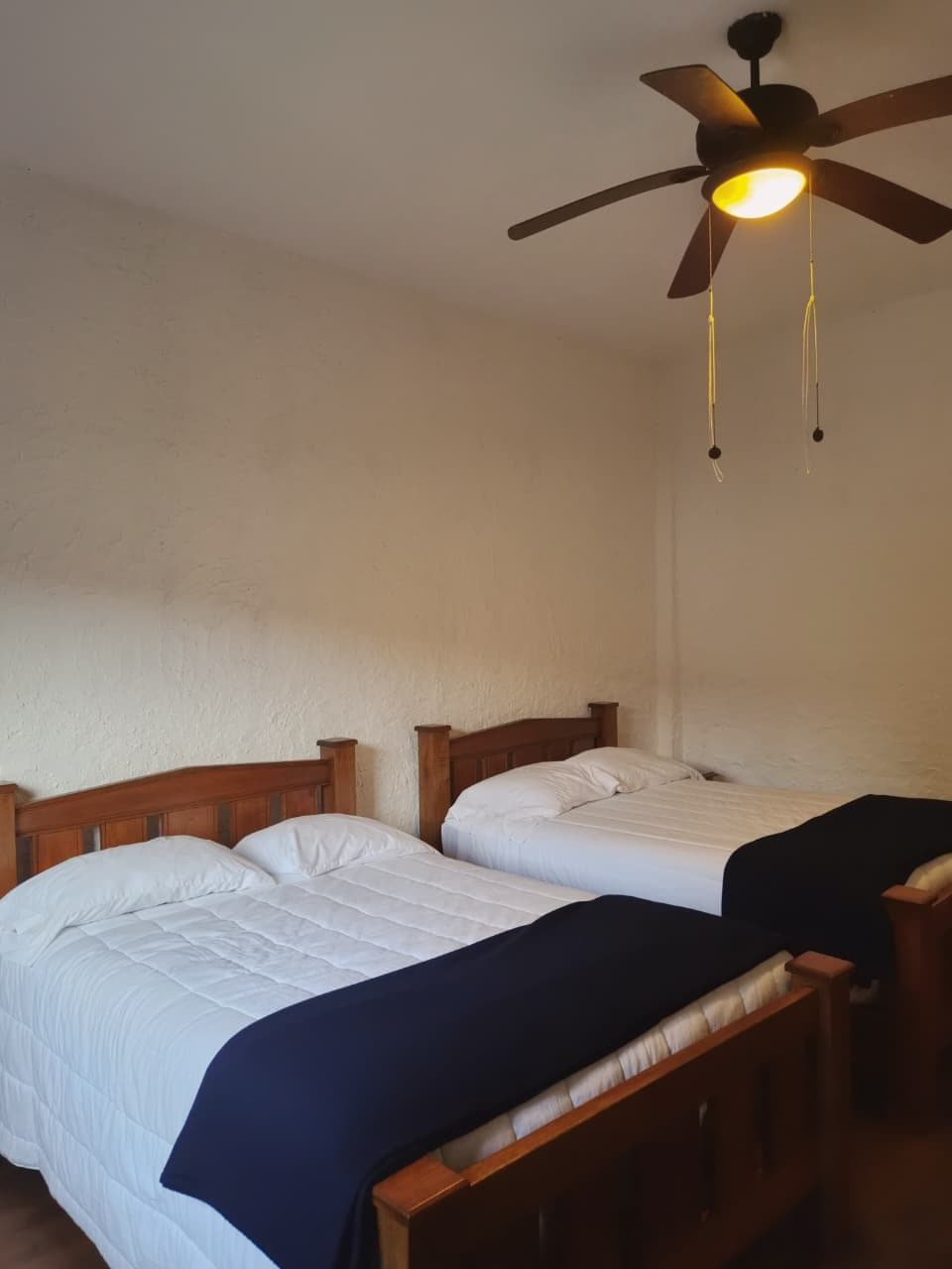 Quarto com duas camas de madeira, um ventilador de teto e um candeeiro.