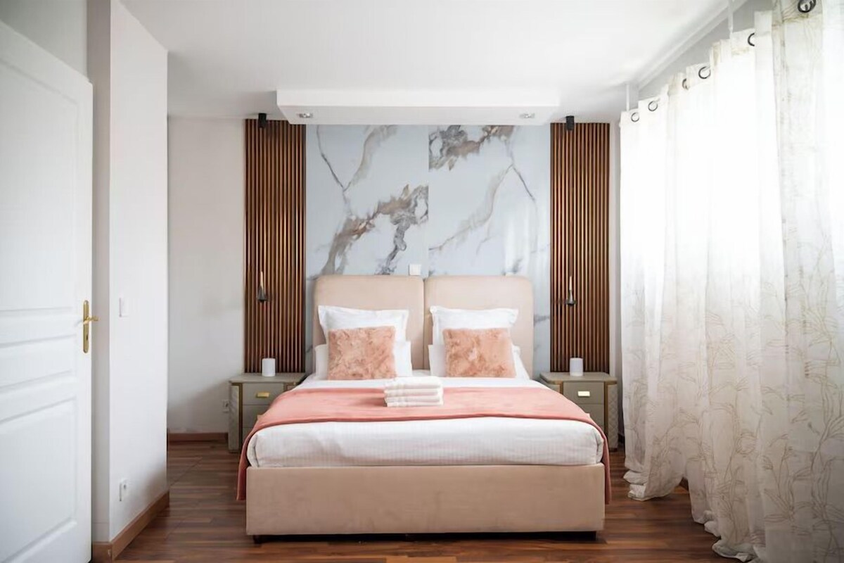 Quarto com cama, duas mesas de cabeceira, cabeceira com design semelhante a mármore, painéis de madeira e janela com cortinas leves.