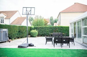 Um quintal com um aro de basquete, uma mesa de pátio com cadeiras e uma janela grande.