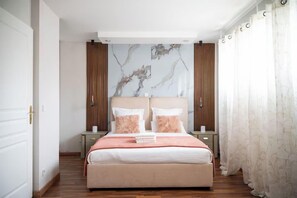 Quarto com cama, duas mesas de cabeceira, cabeceira com design semelhante a mármore, painéis de madeira e janela com cortinas leves.