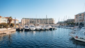 Uma marina com barcos atracados, um edifício grande ao fundo e um céu claro.