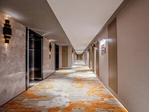 Um corredor de hotel comprido e estreito com carpete estampado, portas de elevador de vidro e luminárias fixadas na parede.