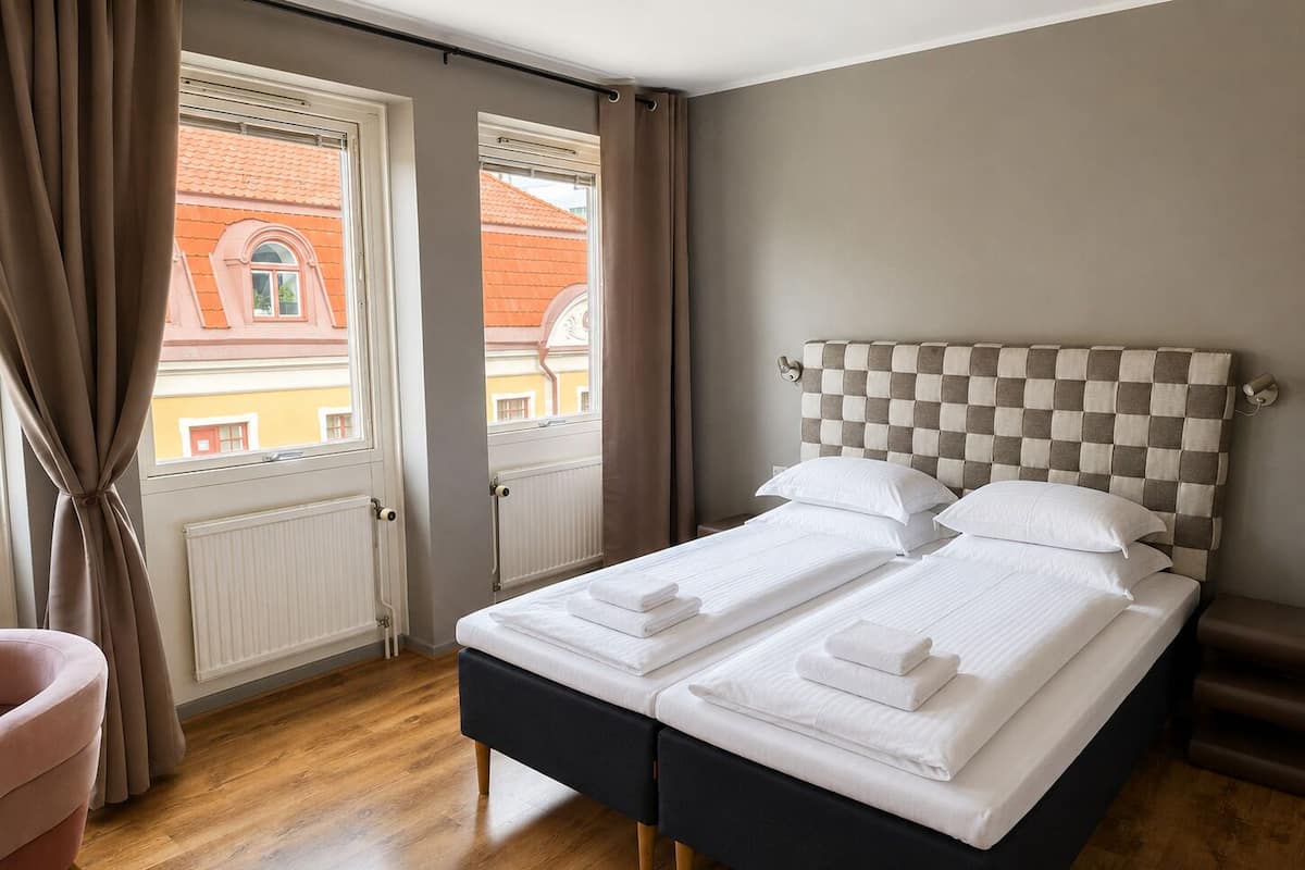Uma cama de casal com roupa de cama e toalhas brancas, cabeceira xadrez, piso de madeira e vista para edifícios pela janela.