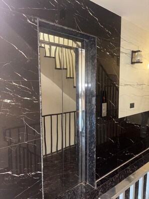Uma escadaria de mármore com porta de elevador de vidro, luminária na parede e corrimão de metal.