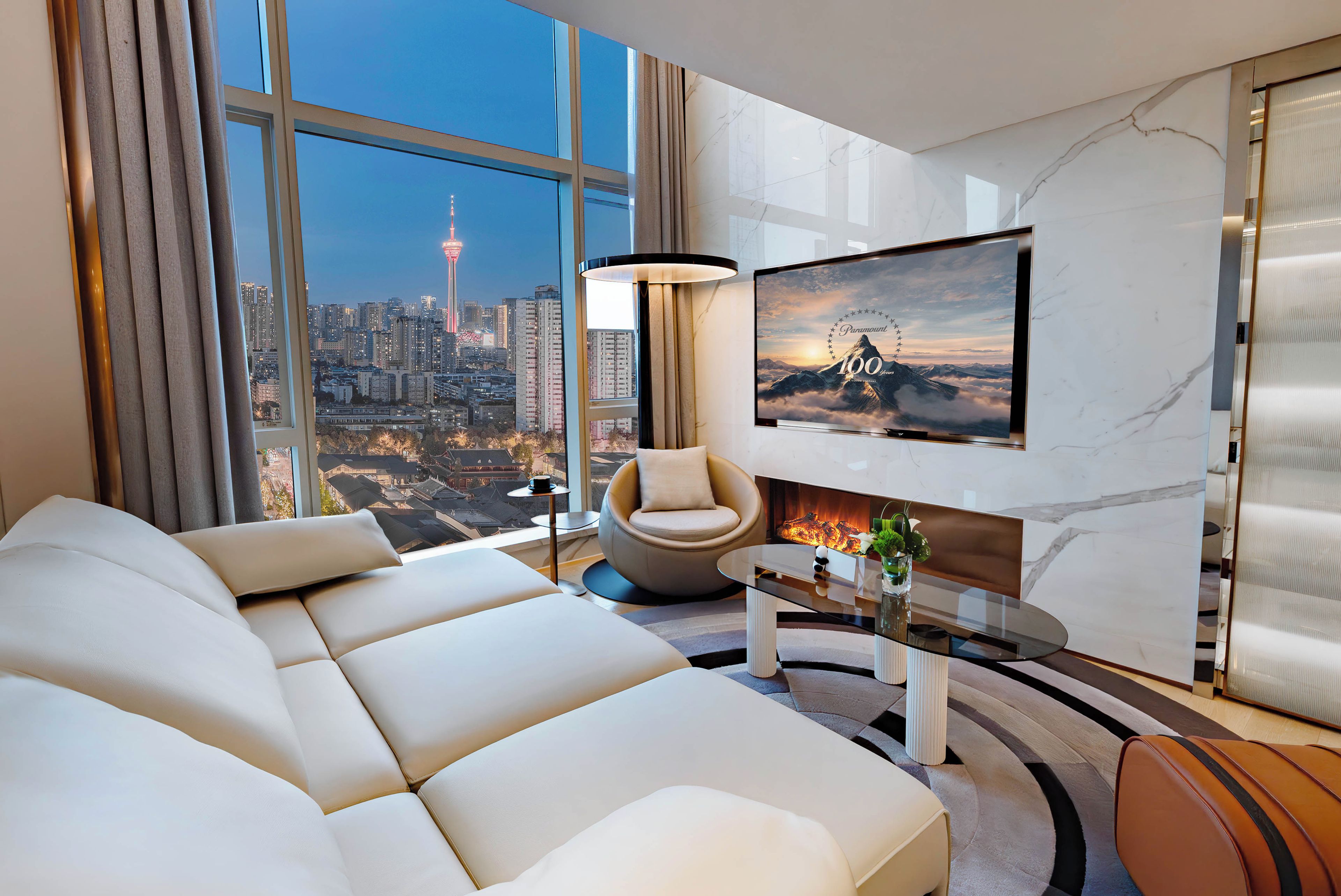 Uma sala de estar moderna com um sofá grande, uma mesa de centro redonda e uma TV na parede. Uma vista para a cidade é visível pela janela.