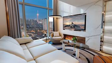 Uma sala de estar moderna com um sofá grande, uma mesa de centro redonda e uma TV na parede. Uma vista para a cidade é visível pela janela.