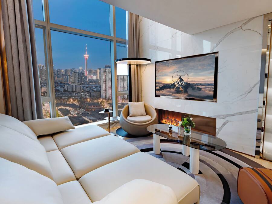 Uma sala de estar moderna com um sofá grande, uma mesa de centro redonda e uma TV na parede. Uma vista para a cidade é visível pela janela.