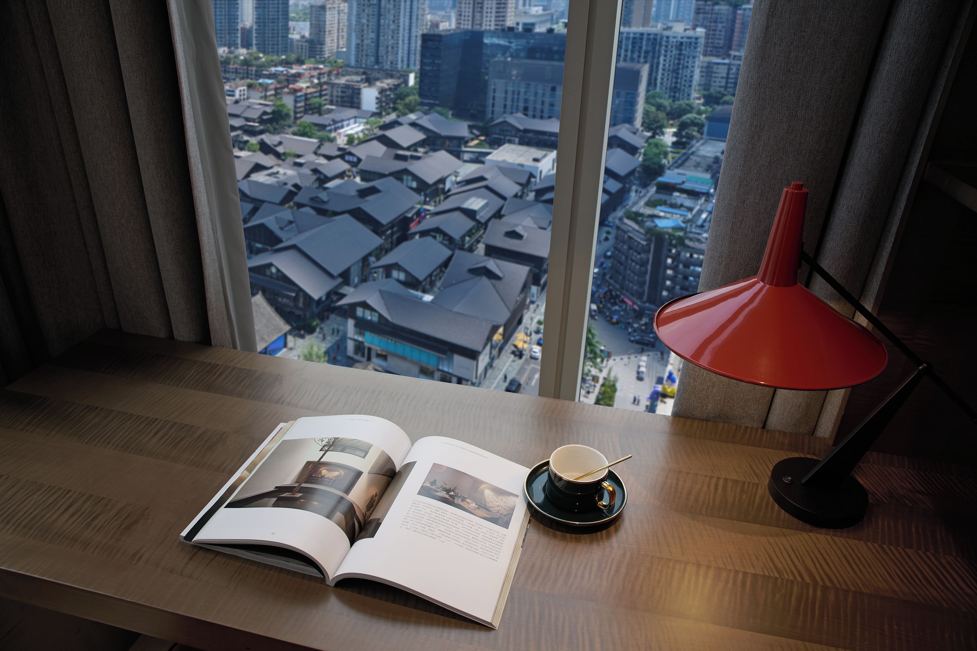 Um livro sobre uma mesa com uma xícara e pires ao lado, um abajur vermelho e vista para uma paisagem urbana através de uma janela.