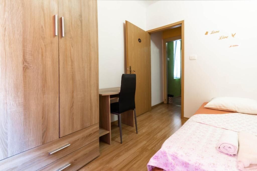 Quarto com guarda-roupa de madeira, escrivaninha com cadeira, cama com colcha rosa e decoração 'Love Live' na parede.