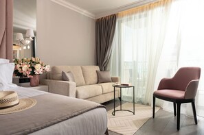 Quarto de hotel com cama, sofá, poltrona, mesa pequena e janela com cortinas.